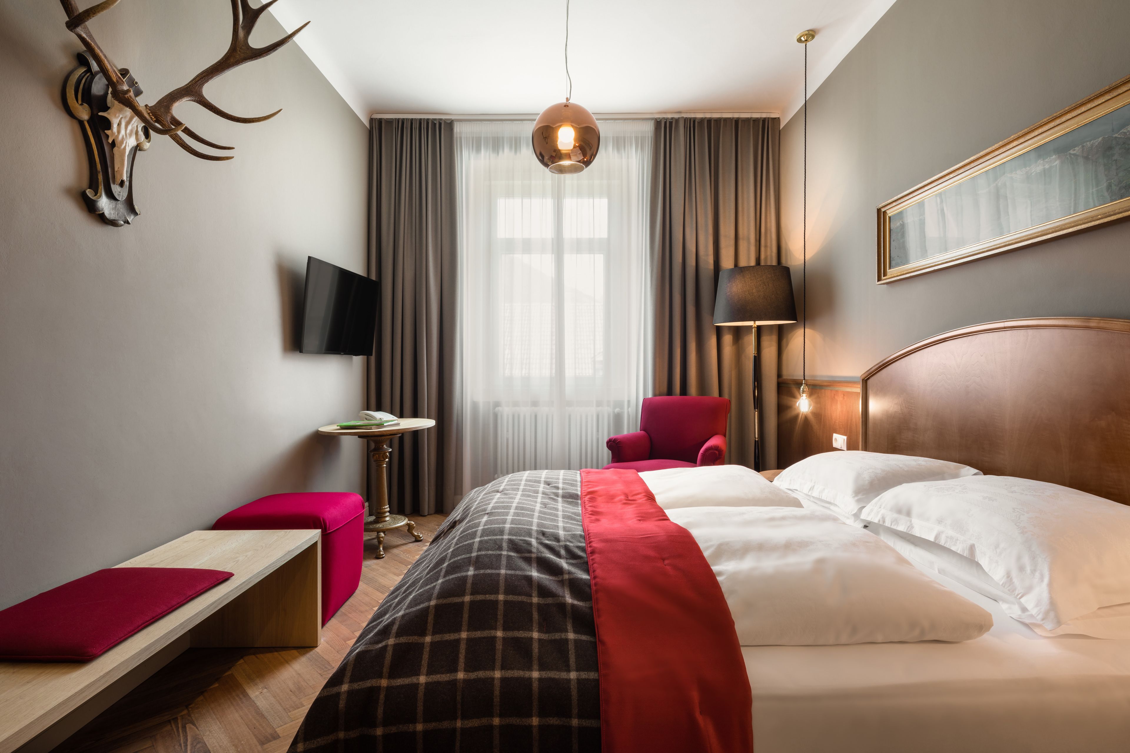 Quarto de hotel com uma cama, uma cadeira vermelha, uma mesinha, cabeça de veado montada, uma televisão e um quadro na parede.