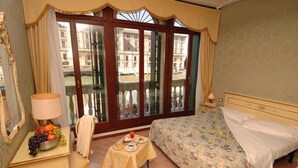 Quarto de hotel com cama, mesa de jantar com frutas, uma cadeira e vista para edifícios através de uma porta de vidro.