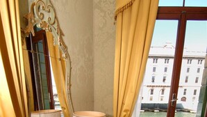 Um quarto com piso de madeira, uma cama, uma mesa com uma fruteira, um abajur, um espelho e uma janela com cortinas.