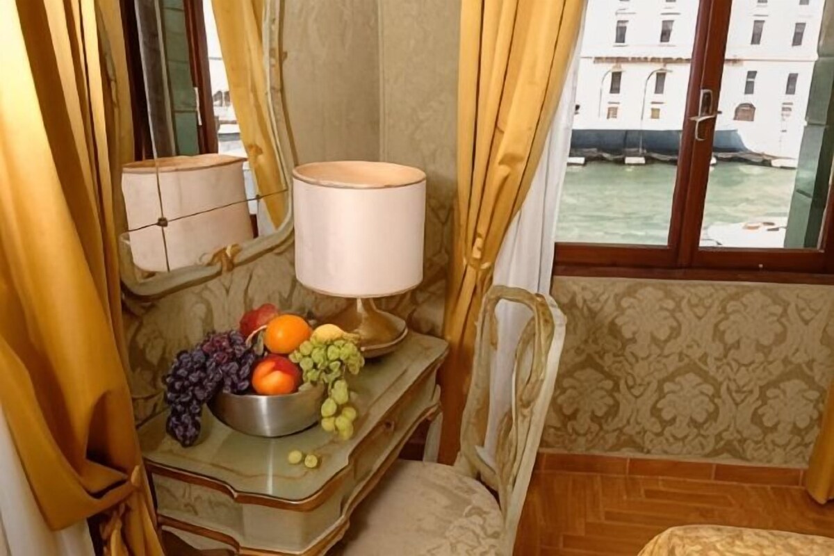 Quarto com uma cama, mesa de cabeceira com abajur, uma tigela de frutas e janela com vista para edifícios e água.