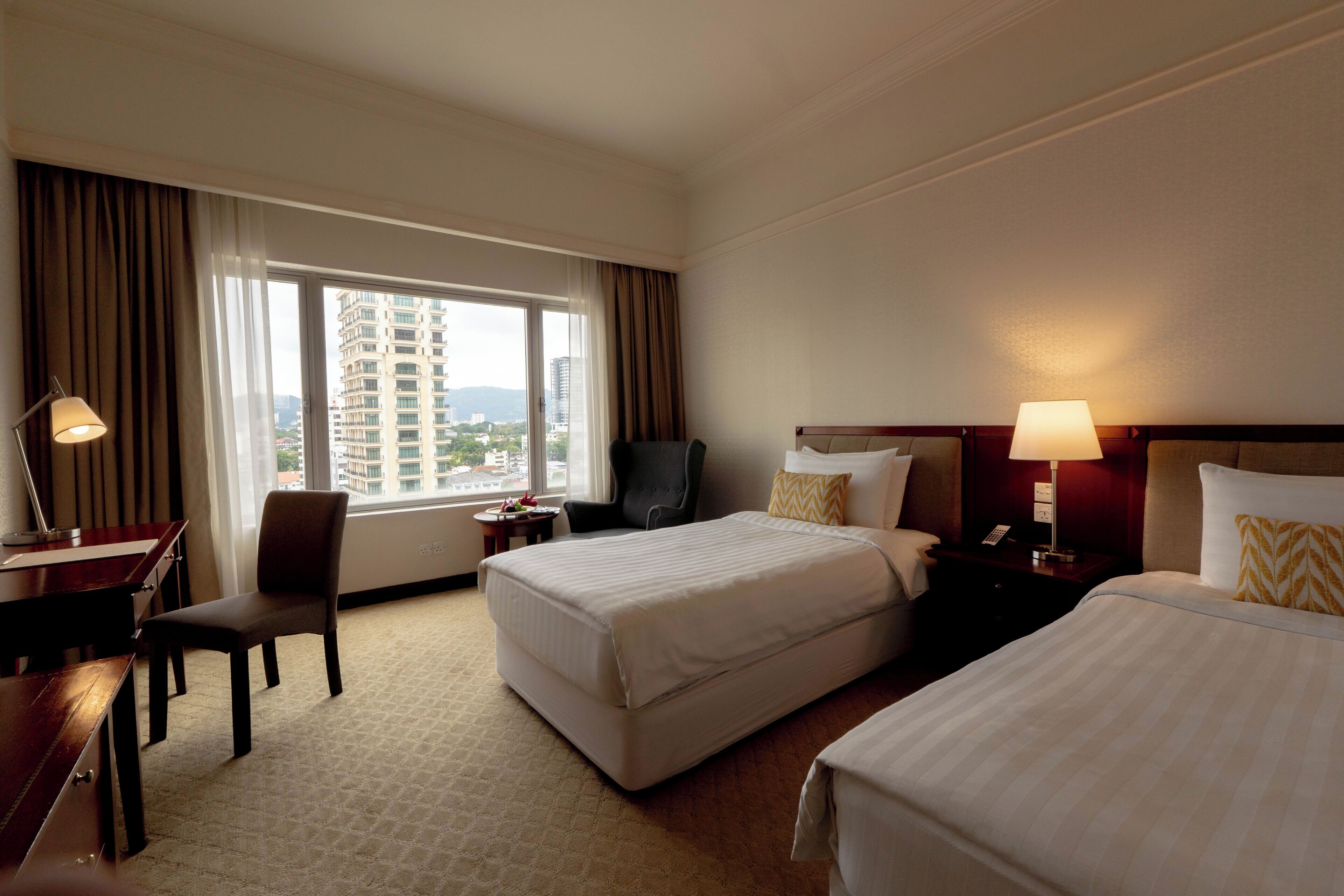 Quarto de hotel com duas camas, uma escrivaninha, uma cadeira, um abajur e vista para a paisagem urbana.