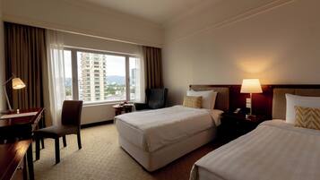 Quarto de hotel com duas camas, uma escrivaninha, uma cadeira, um abajur e vista para a paisagem urbana.