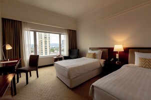 Quarto de hotel com duas camas, uma escrivaninha, uma cadeira, um abajur e vista para a paisagem urbana.