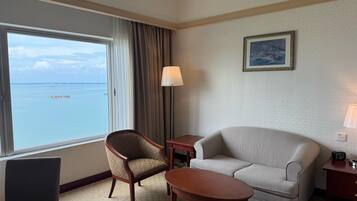 Quarto de hotel com uma janela grande que oferece vista para o mar, um sofá, uma cadeira, uma mesa e um abajur.