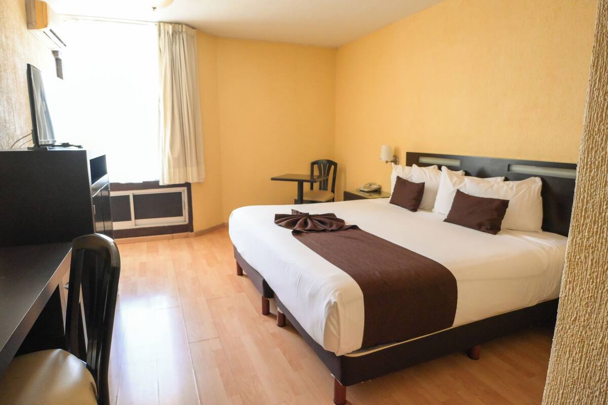 Quarto de hotel com cama, escrivaninha, cadeira, mesa pequena e janela com cortinas.