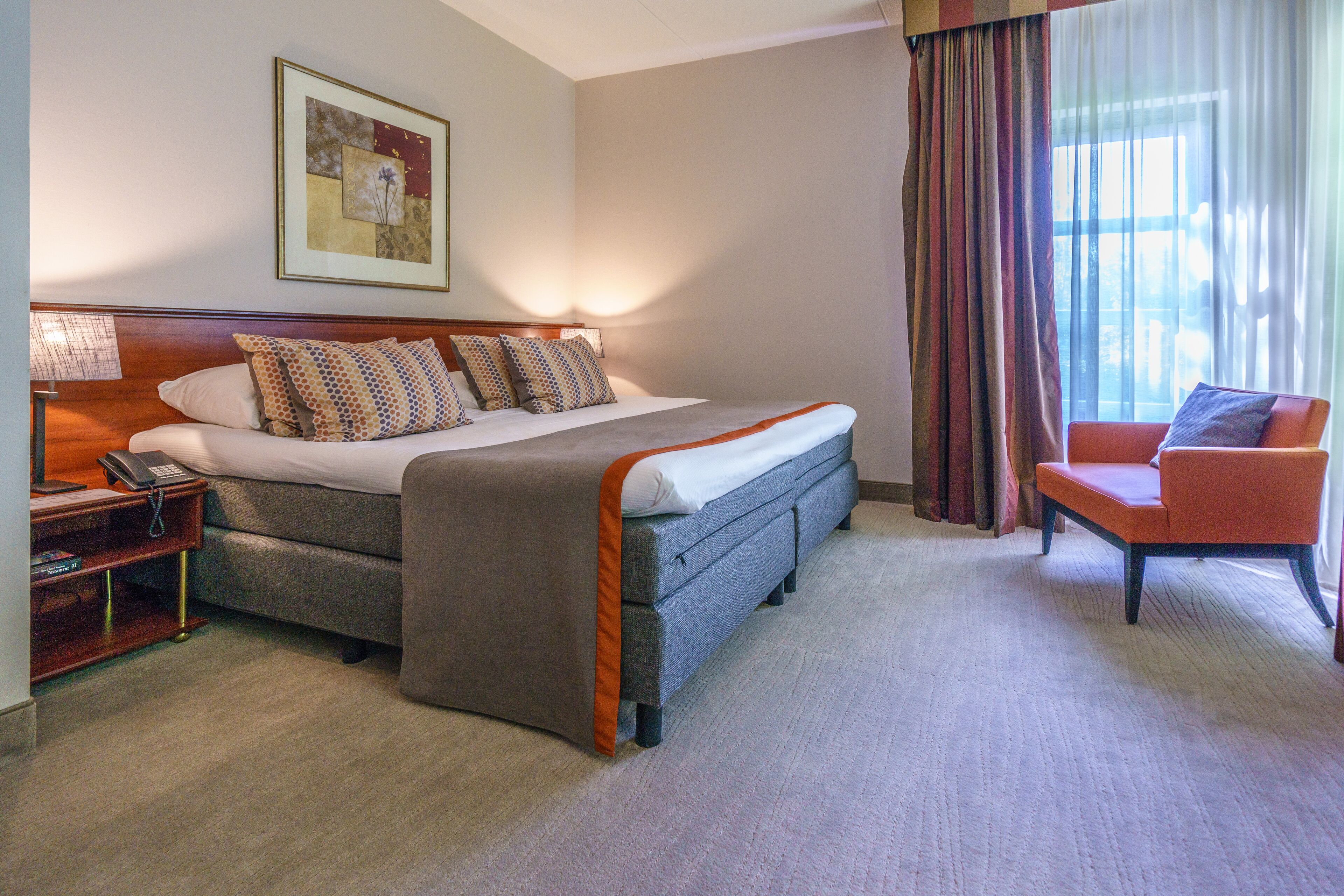 Quarto de hotel com uma cama, uma cadeira, uma escrivaninha com abajur e um quadro na parede.