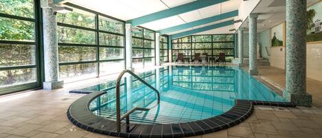 Piscina interior com paredes de vidro transparente, piso ladrilhado e algumas cadeiras de descanso.