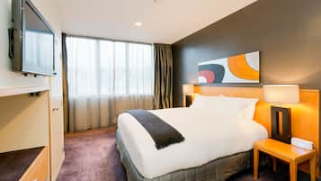 Quarto de hotel com cama, televisão na parede, mesa de cabeceira com abajur e janela com cortinas.
