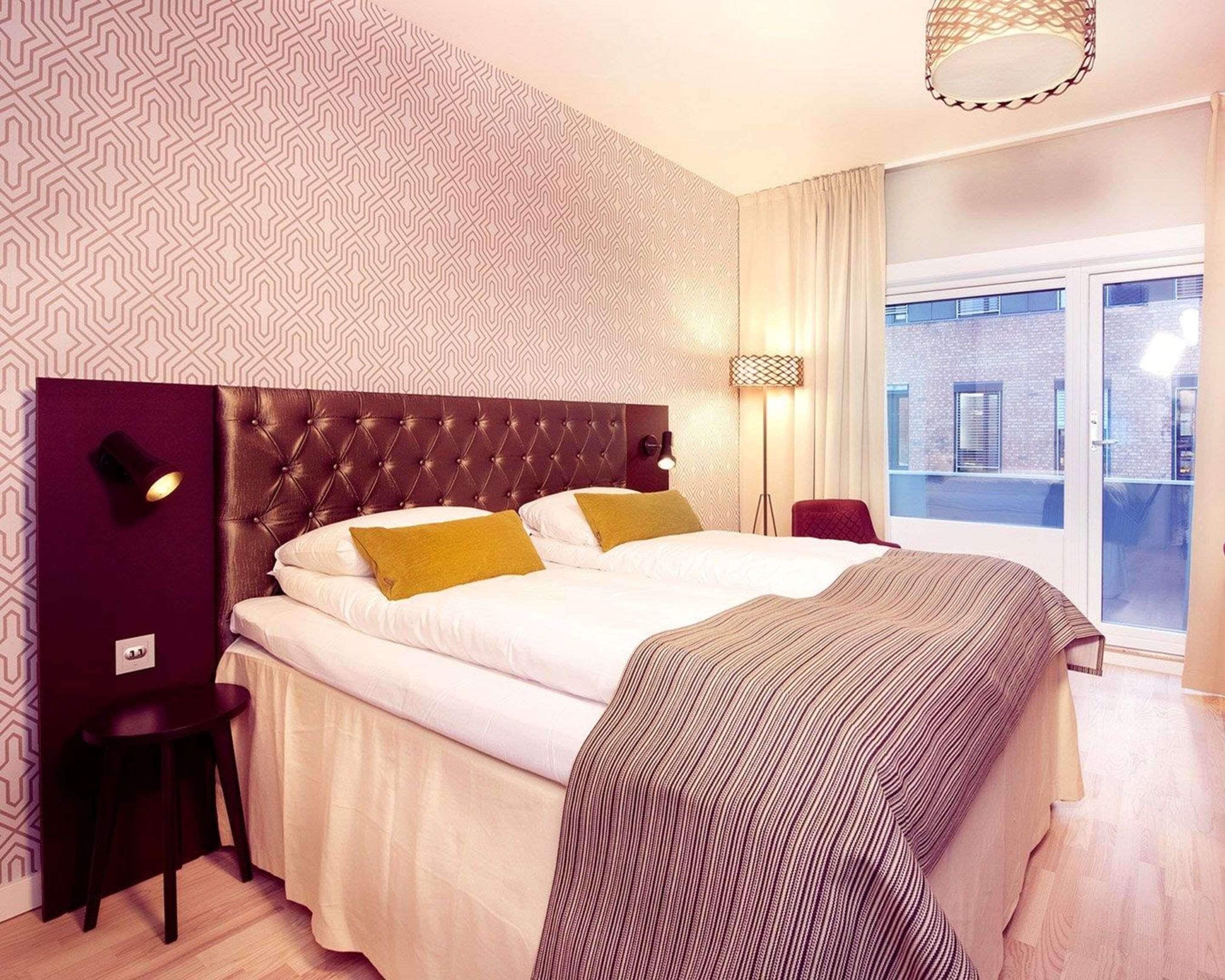 Quarto de hotel moderno com uma cama grande, cabeceira, mesa de cabeceira, abajur e vista da cidade através de uma janela ampla.