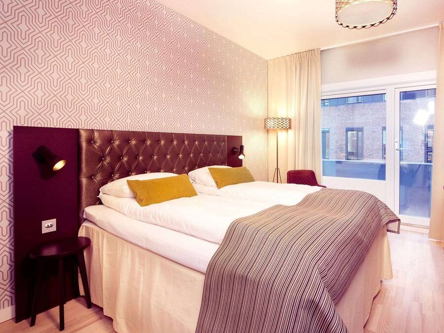 Quarto de hotel moderno com uma cama grande, cabeceira, mesa de cabeceira, abajur e vista da cidade através de uma janela ampla.