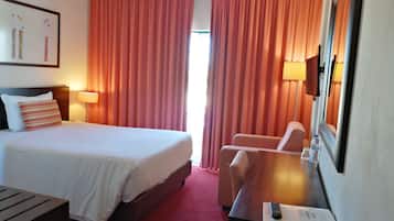 Quarto de hotel com uma cama, escrivaninha, cadeira, abajur e televisão.