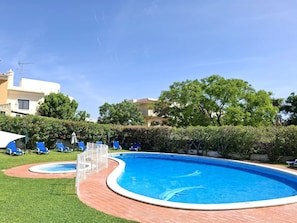 Piscina com borda curva, rodeada por um deck de tijolos e cadeiras de sol azuis.