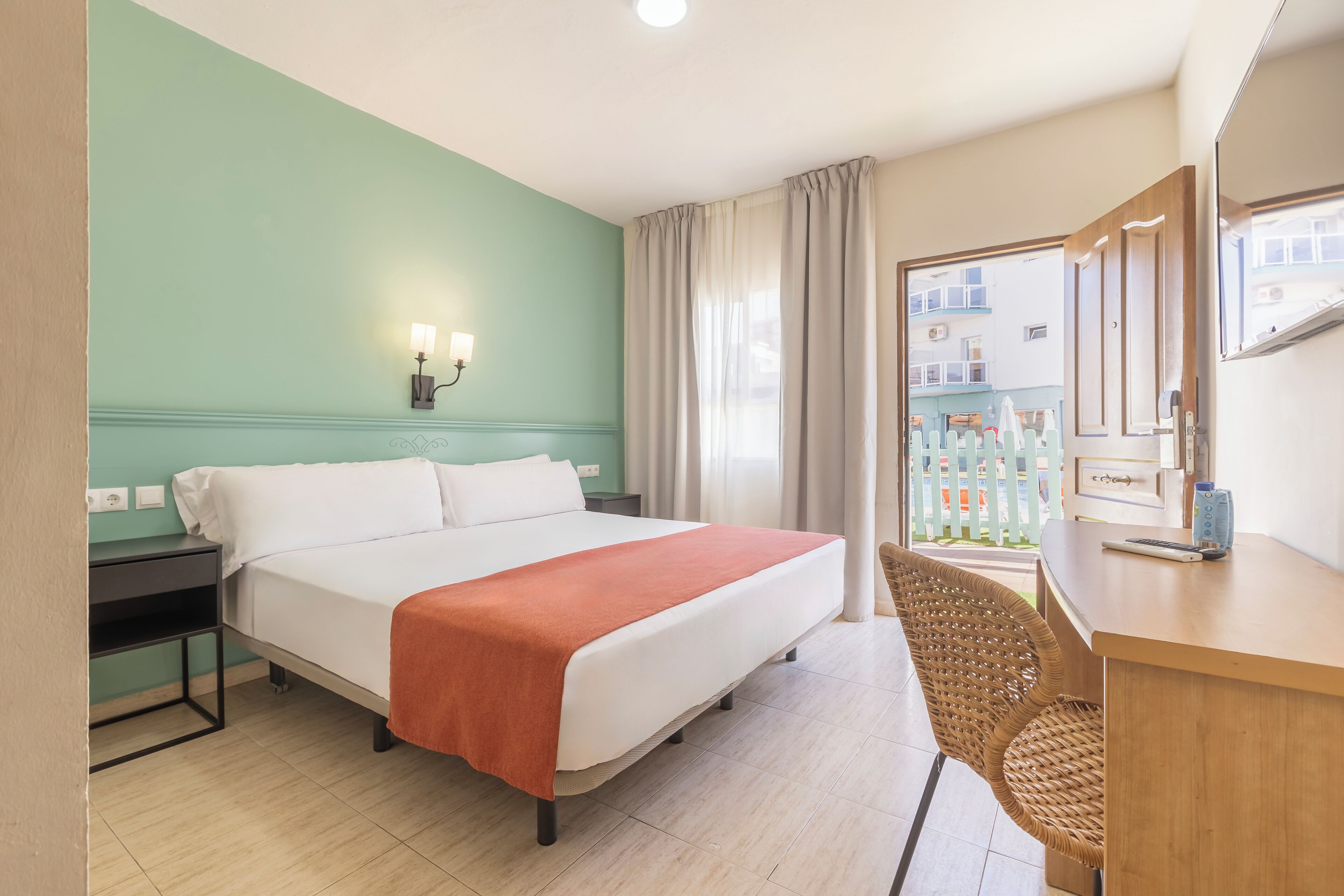 Quarto de hotel com cama, escrivaninha, cadeira, janela com cortinas e luminária na parede.