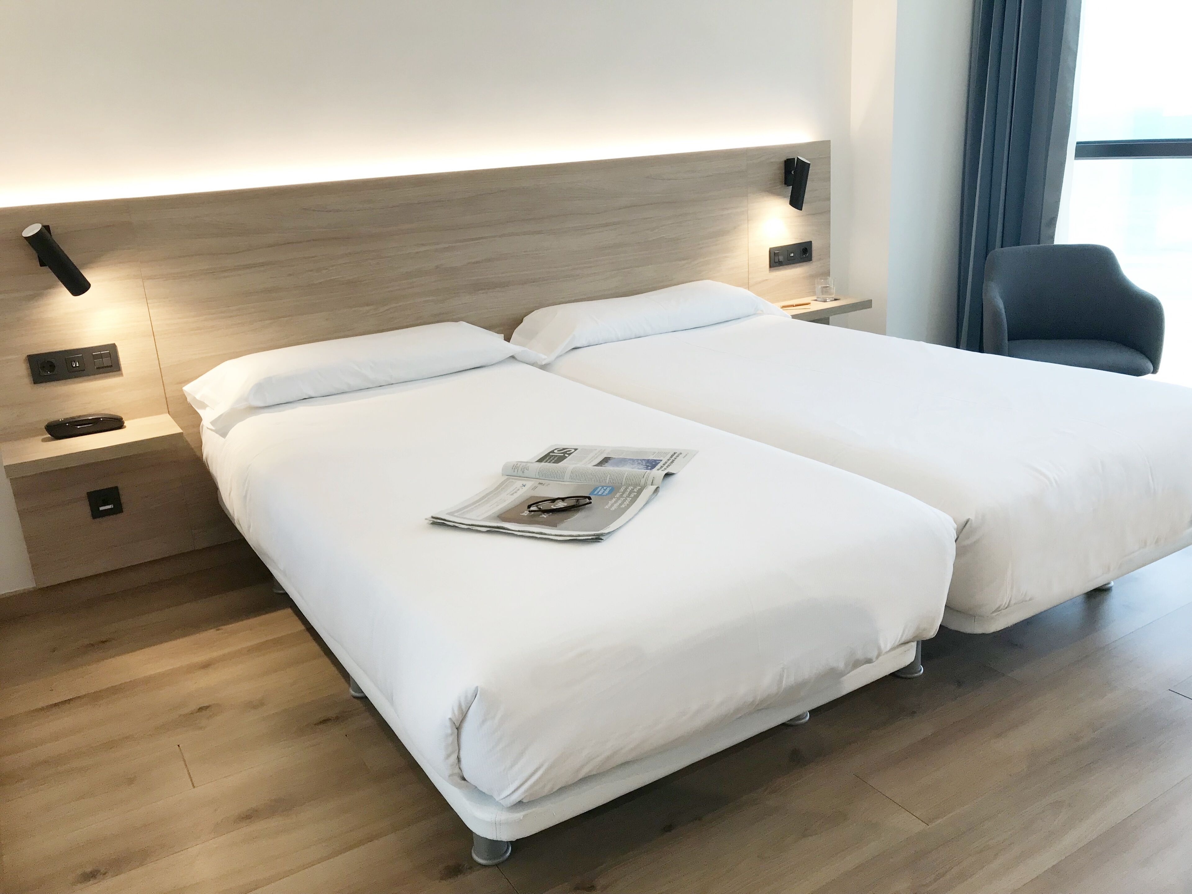 Uma cama de casal com lençóis brancos, uma mesa de cabeceira com abajur e uma revista sobre a cama.