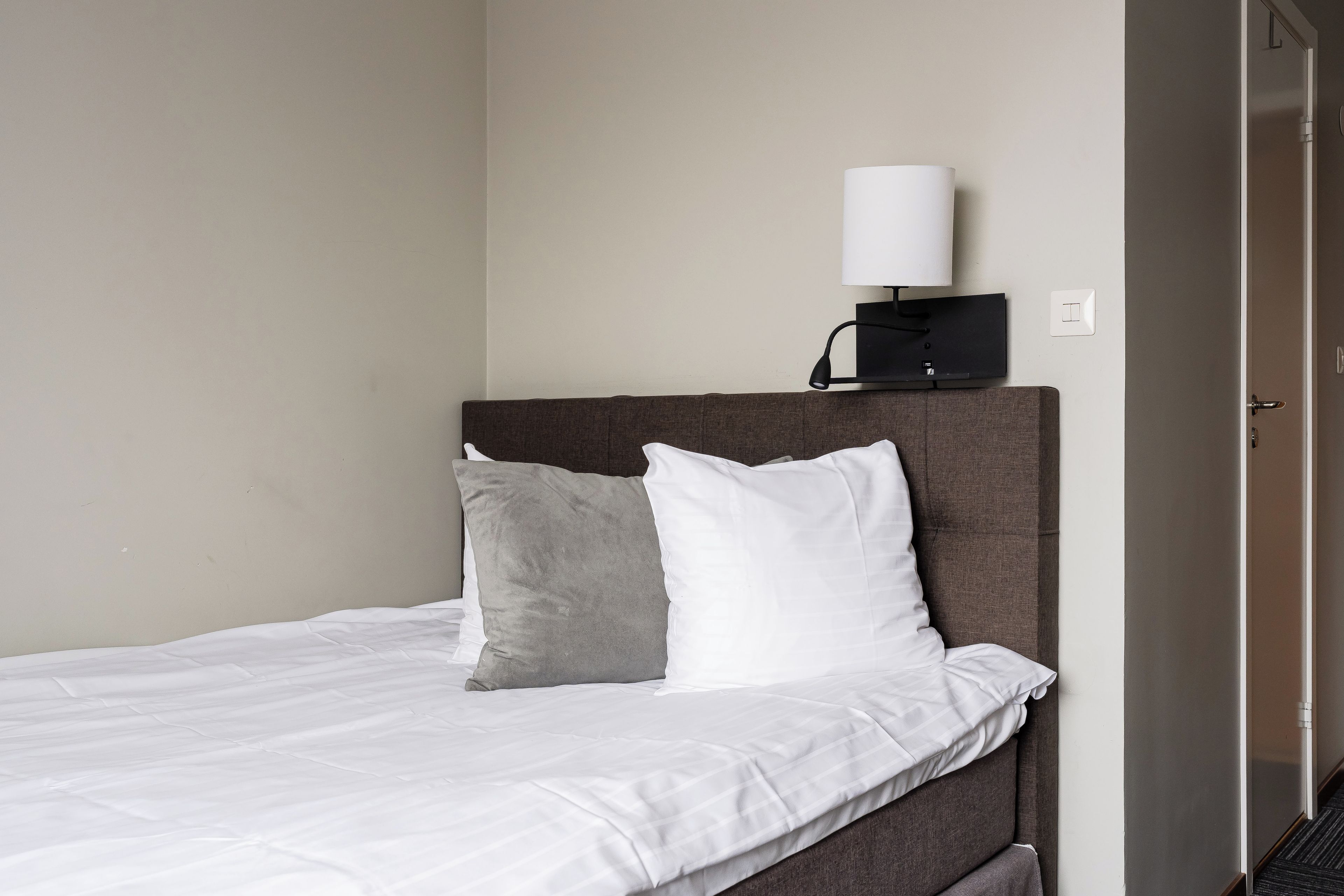 Uma cama de solteiro com roupa de cama branca, um travesseiro cinzento e uma luminária de cabeceira.