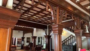 Um hall espaçoso com altos tetos de madeira e trabalhos em madeira detalhados. Há uma escadaria que leva a um andar superior e um balcão de recepção com um computador e uma placa vermelha.