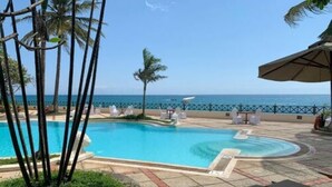 Área de piscina com vista para o oceano, cercada por palmeiras e espreguiçadeiras.
