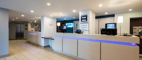 Uma área de recepção de hotel, com balcão de atendimento, relógio e tela digital exibindo informações.
