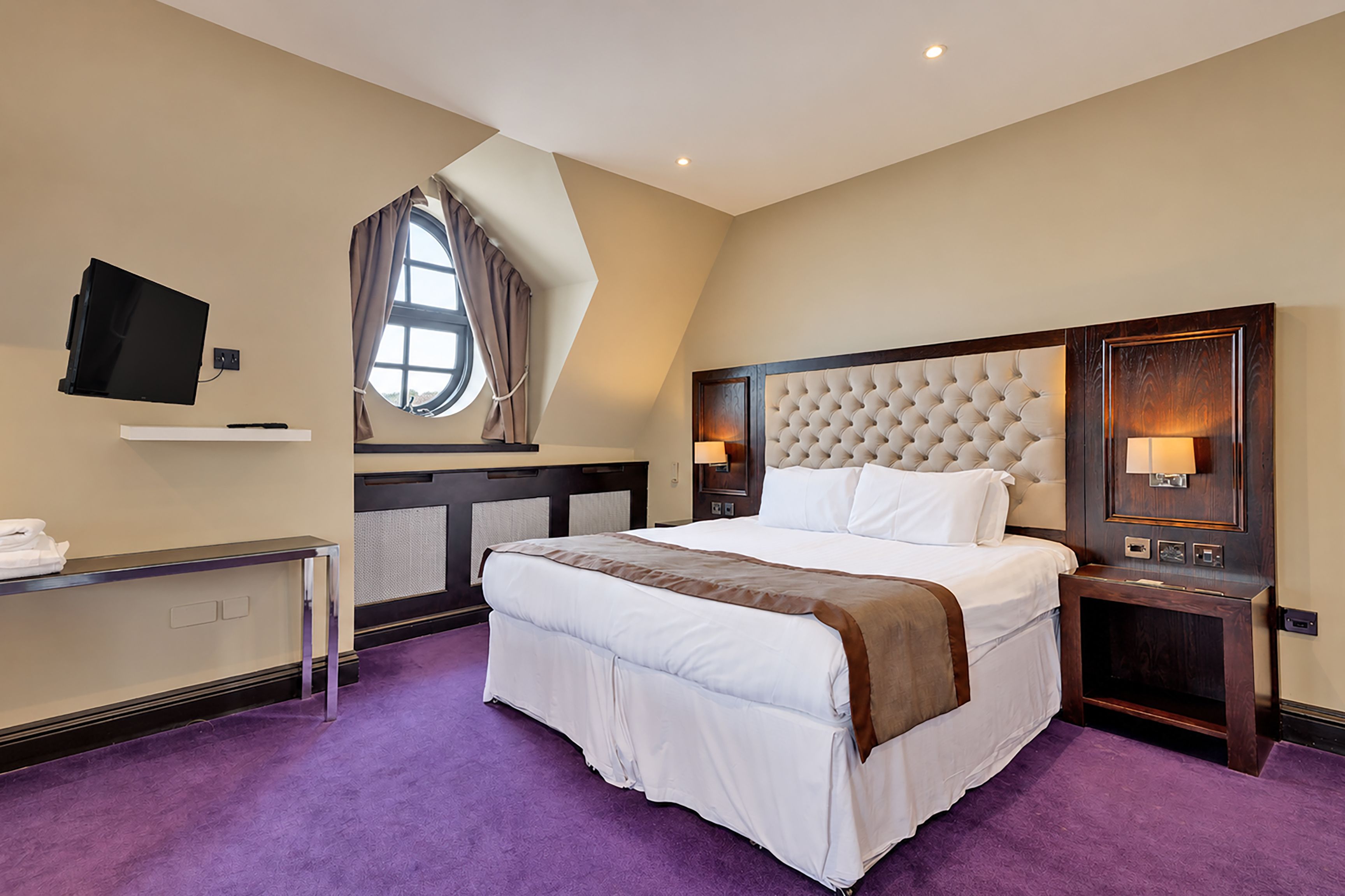 Quarto de hotel com uma cama grande, uma escrivaninha, TV de tela plana, janela com cortinas e mesa de cabeceira com abajur.