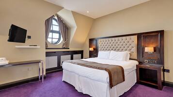 Quarto de hotel com uma cama grande, uma escrivaninha, TV de tela plana, janela com cortinas e mesa de cabeceira com abajur.