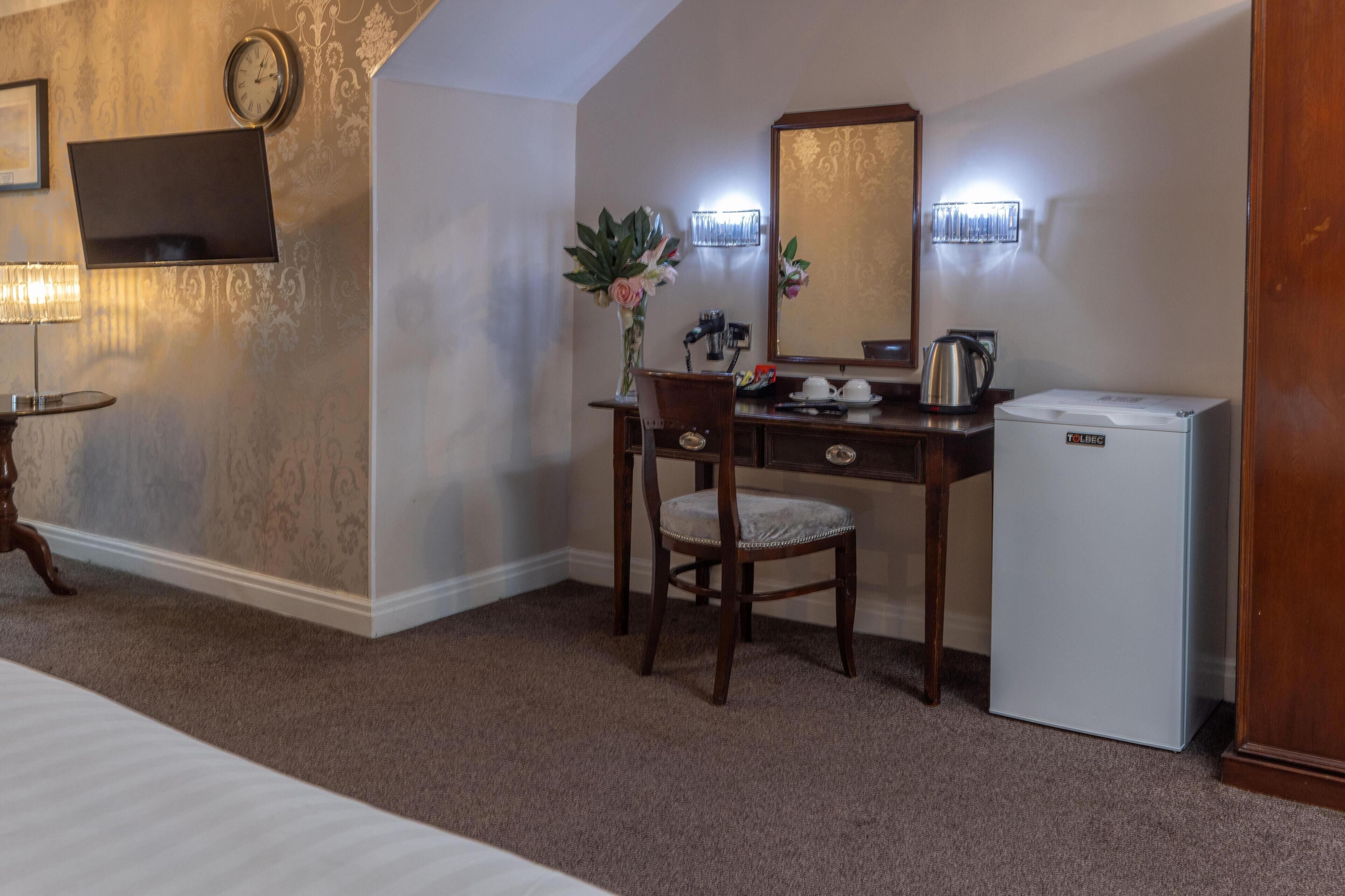 Quarto de hotel com cama, secretária com cadeira, pequeno frigorífico, relógio e televisão fixada na parede.