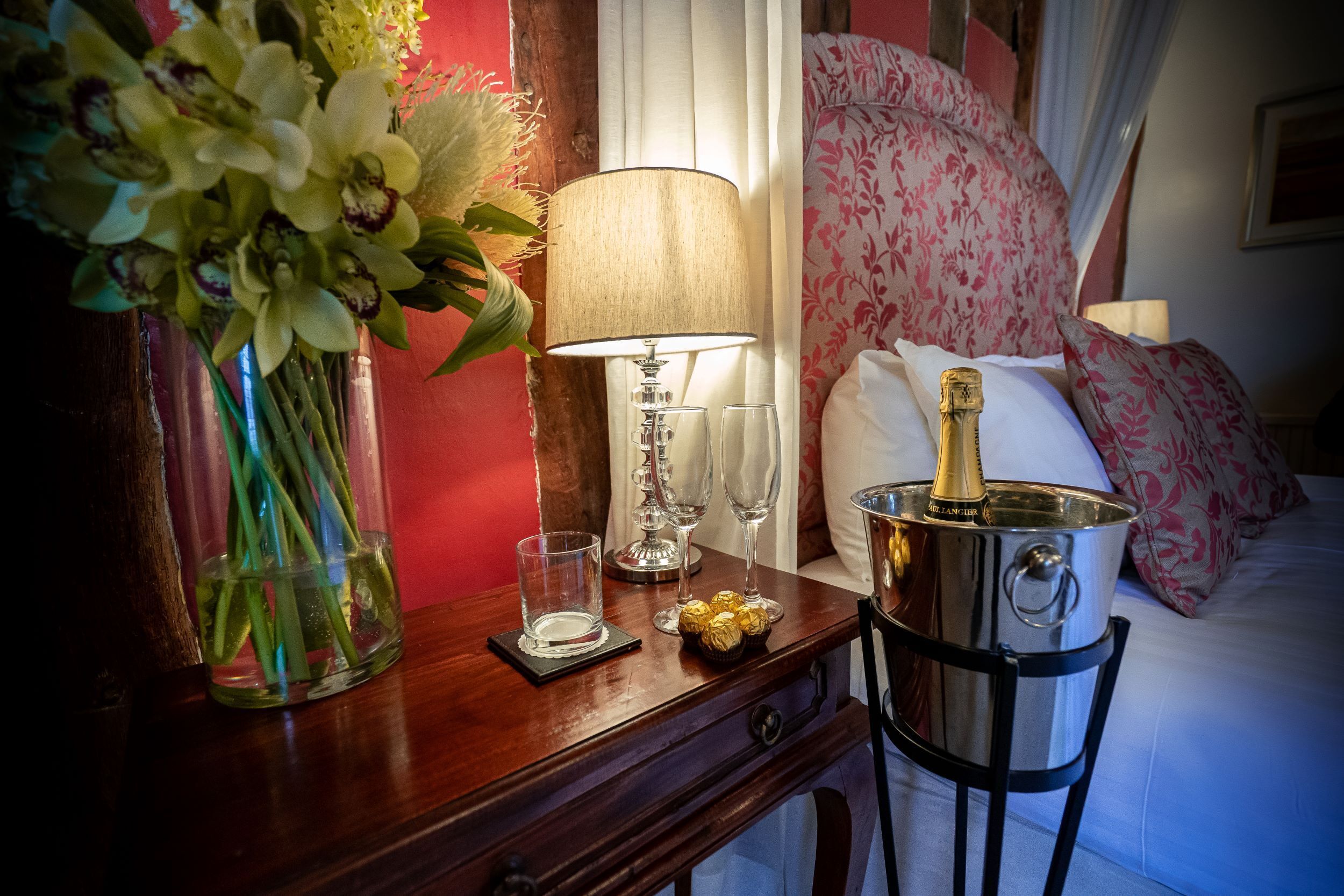 Quarto de hotel com mesa de cabeceira de madeira, um vaso de flores, abajur, uma garrafa de champanhe e uma cama com roupa de cama florida.