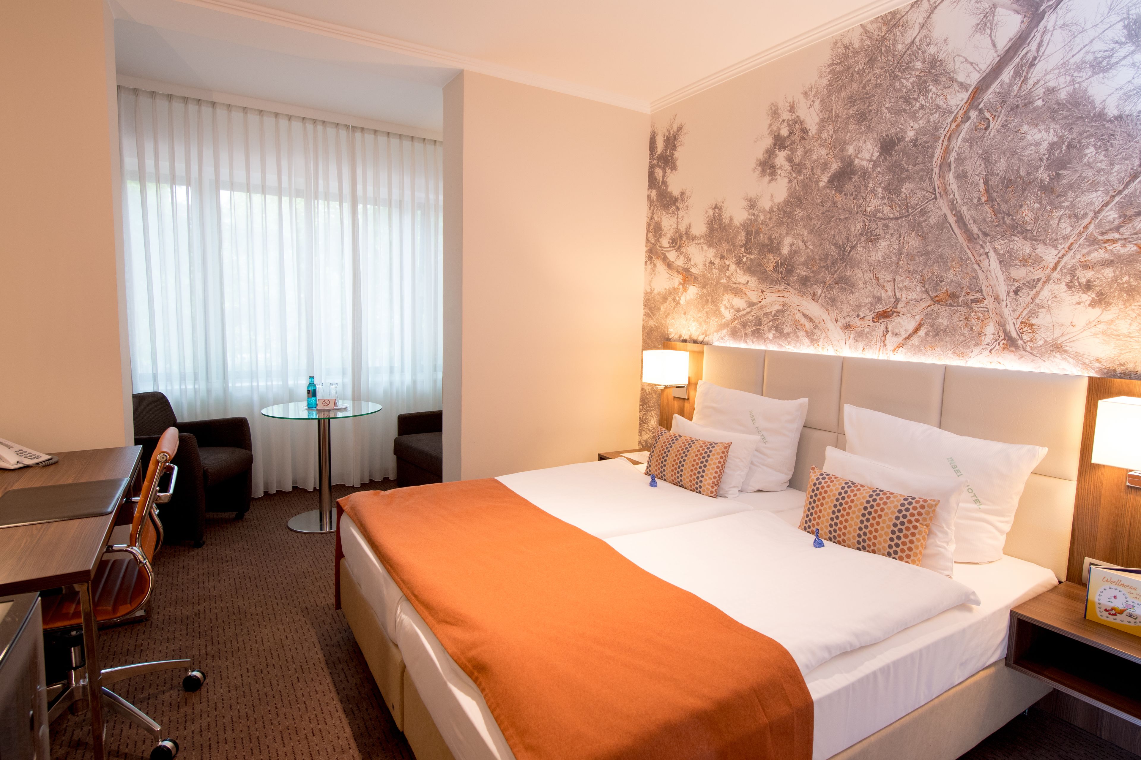 Quarto de hotel com cama grande, escrivaninha, cadeira, mesa pequena e janela com cortinas finas.
