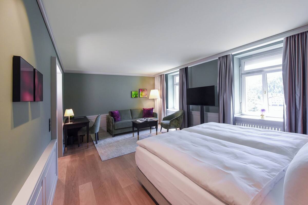 Quarto de hotel com cama grande, escrivaninha com luminária, sofá e uma poltrona. Há uma janela com cortinas e vista para o exterior.