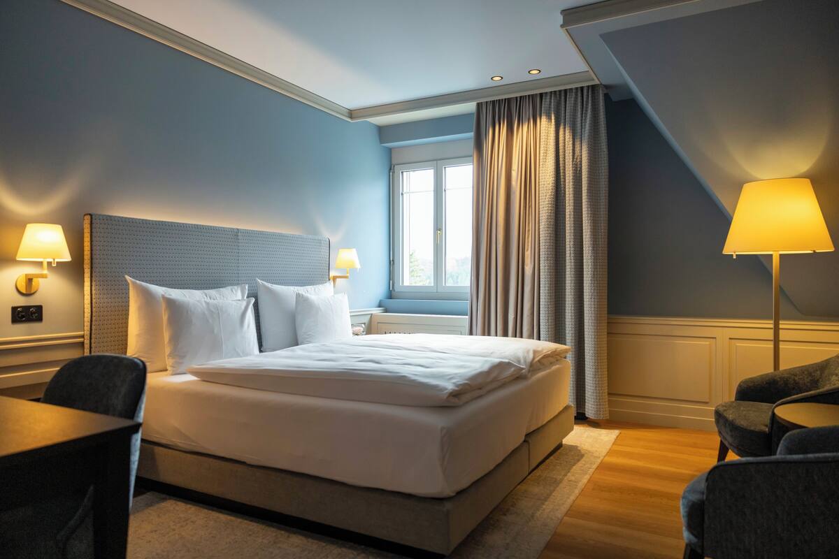 Quarto de hotel com cama, duas mesas de cabeceira, escrivaninha, cadeira, janela com cortinas e luminária.