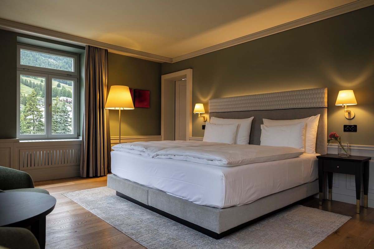 Quarto de hotel com uma cama grande, duas luminárias de cabeceira, uma janela com cortinas, uma mesinha e uma cadeira.