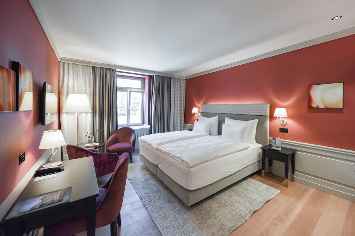 Quarto de hotel com uma cama grande, uma mesa com cadeira, uma mesinha de apoio com abajur e uma janela com cortinas.