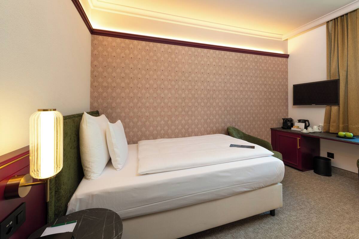 Quarto de hotel com cama, secretária, televisão e TV fixada na parede.