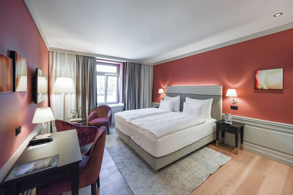 Quarto de hotel com uma cama grande, uma mesa com cadeira, uma mesinha de apoio com abajur e uma janela com cortinas.