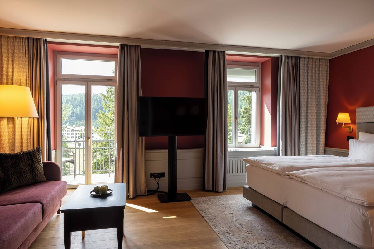 Quarto de hotel com cama, televisão, sofá, mesa e janela com cortinas.