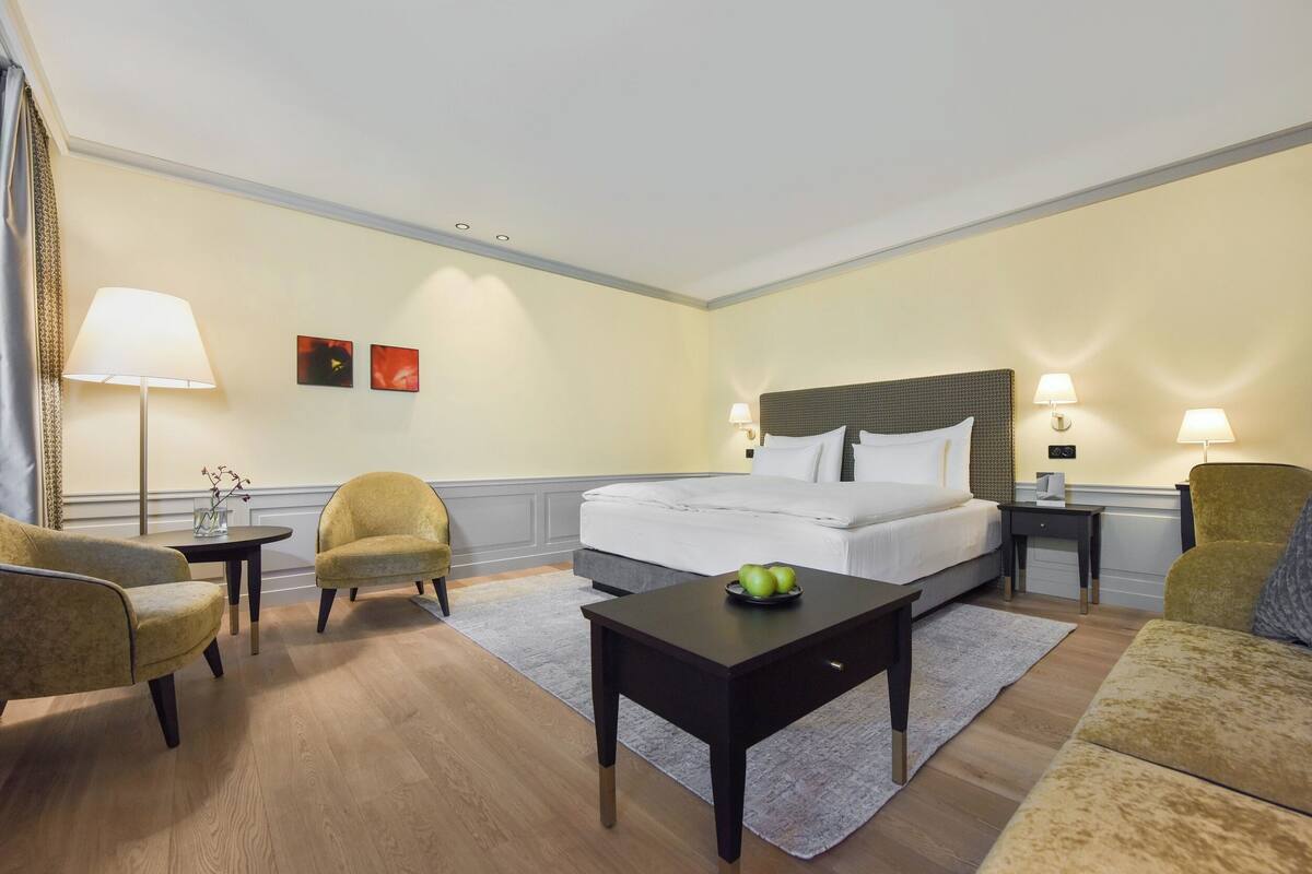 Quarto de hotel com uma cama, duas poltronas, uma mesa pequena e uma mesinha de cabeceira.