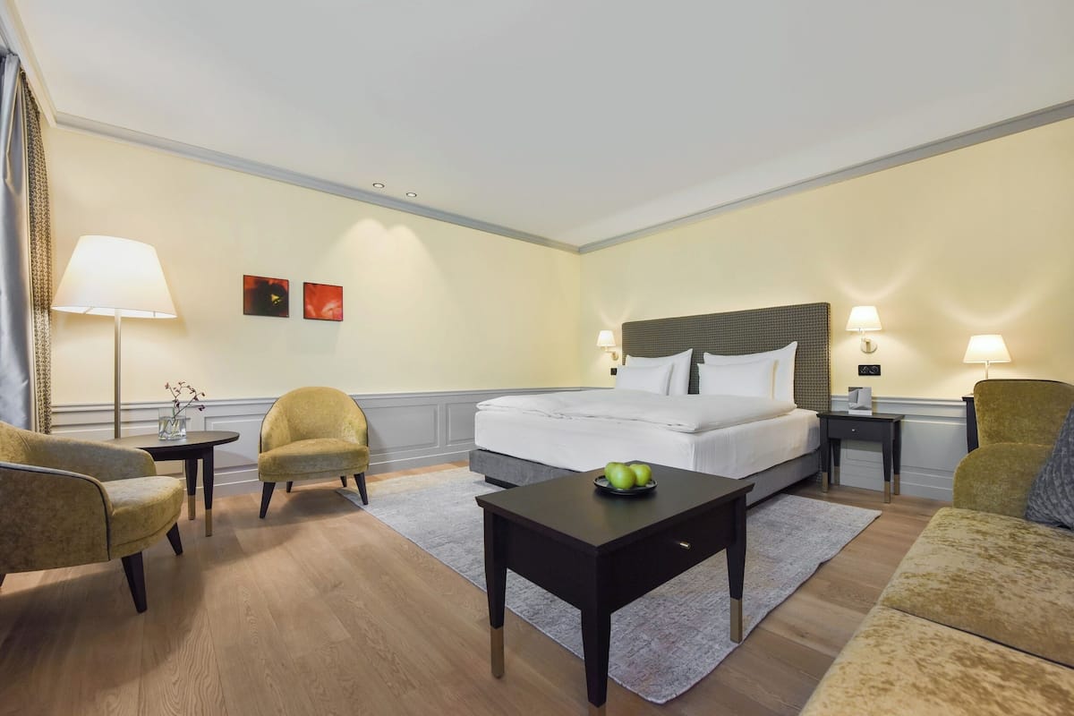 Quarto de hotel com uma cama, duas poltronas, uma mesa pequena e uma mesinha de cabeceira.