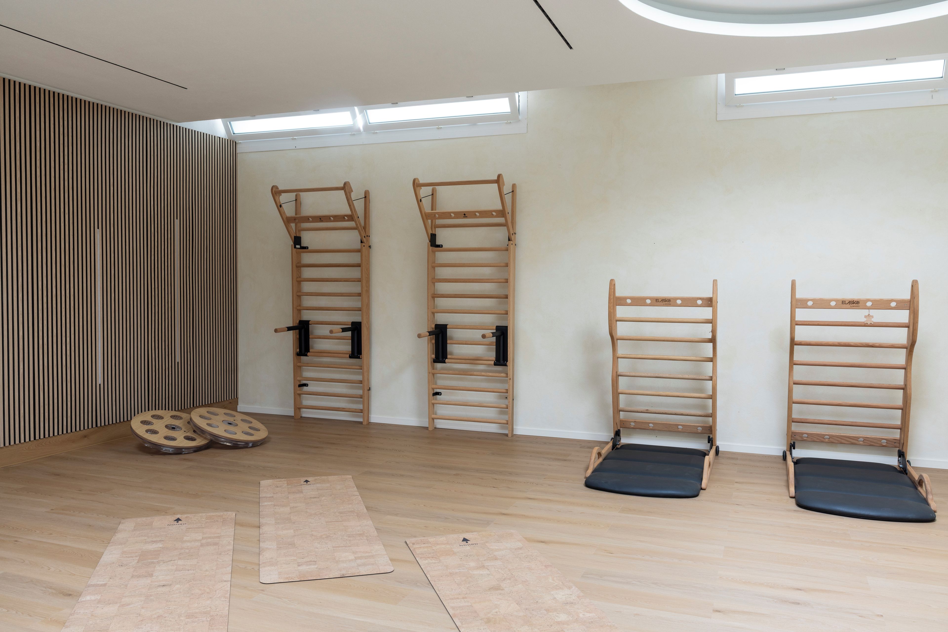 Quarto com teto inclinado, piso de madeira e três equipamentos de exercício fixados na parede.