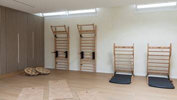 Quarto com teto inclinado, piso de madeira e três equipamentos de exercício fixados na parede.