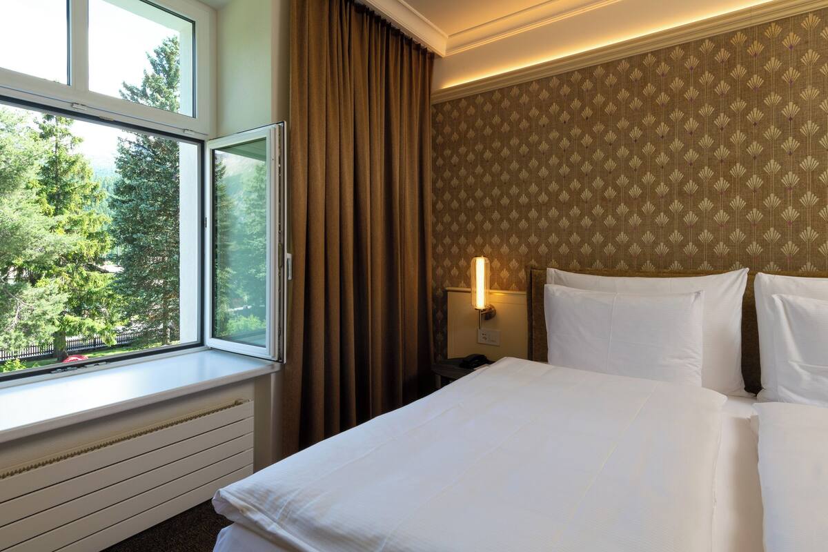 Quarto de hotel com uma janela grande, uma cama com roupa de cama branca, uma mesa de cabeceira e uma parede com papel de parede estampado.