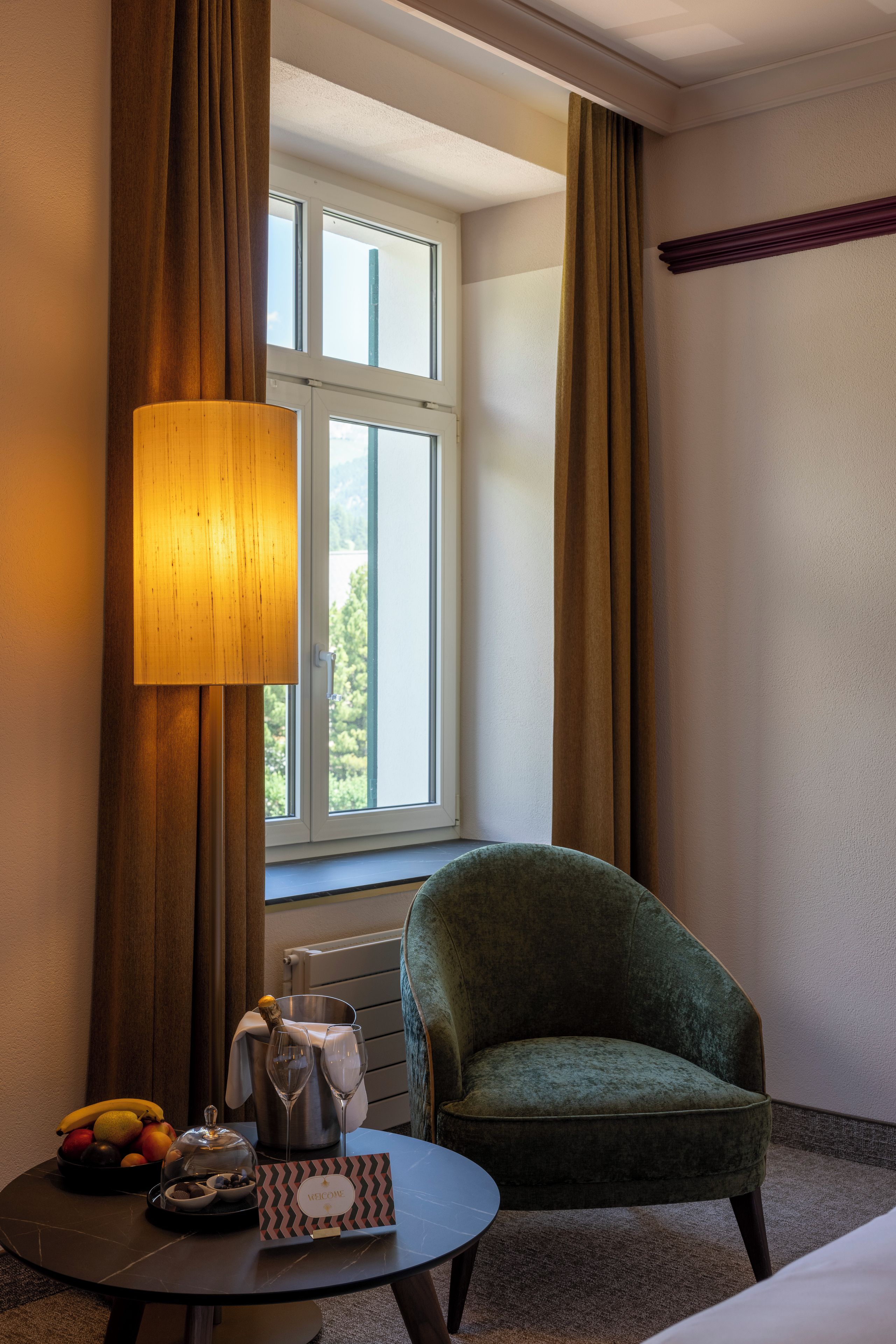 Quarto de hotel com uma poltrona verde, uma mesa redonda pequena com uma fruteira e um copo, e uma janela com cortinas.