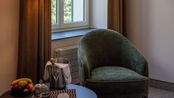 Quarto de hotel com uma poltrona verde, uma mesa redonda pequena com uma fruteira e um copo, e uma janela com cortinas.