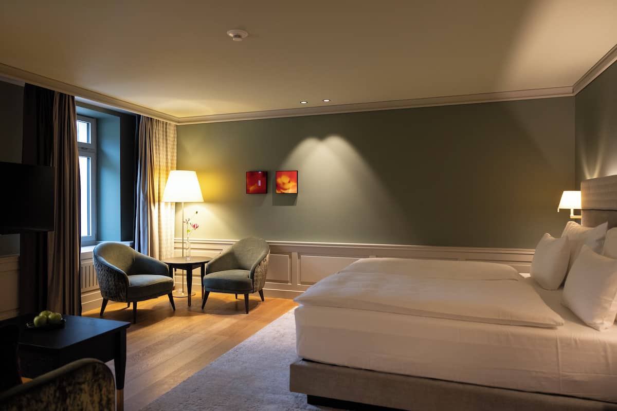 Quarto de hotel com uma cama grande, duas poltronas, uma mesinha e um abajur.