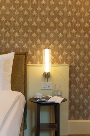 Quarto de hotel com uma cama, uma mesa de cabeceira com uma lâmpada, uma xícara, um livro e uma garrafa.