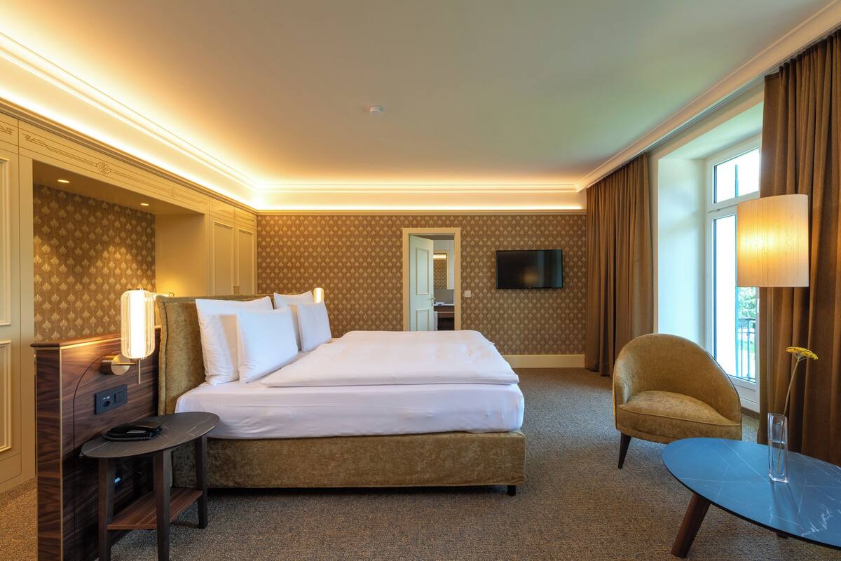 Quarto de hotel com uma cama grande, uma cadeira, uma mesinha, uma televisão e uma janela com cortinas.