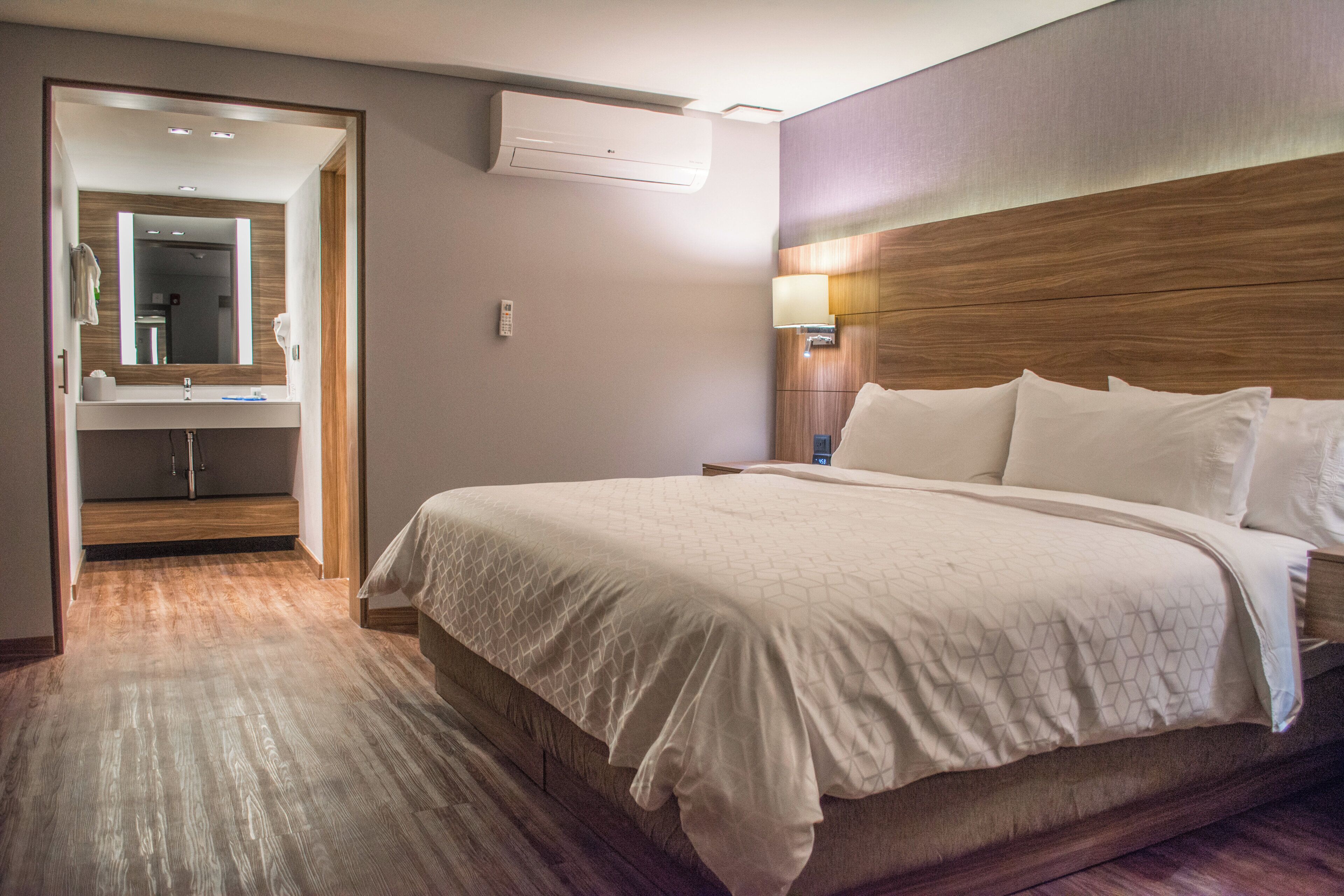 Quarto de hotel com cama, mesa de cabeceira, luminária, ar condicionado, banheiro com espelho e porta-toalhas.