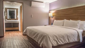 Quarto de hotel com cama, mesa de cabeceira, luminária, ar condicionado, banheiro com espelho e porta-toalhas.