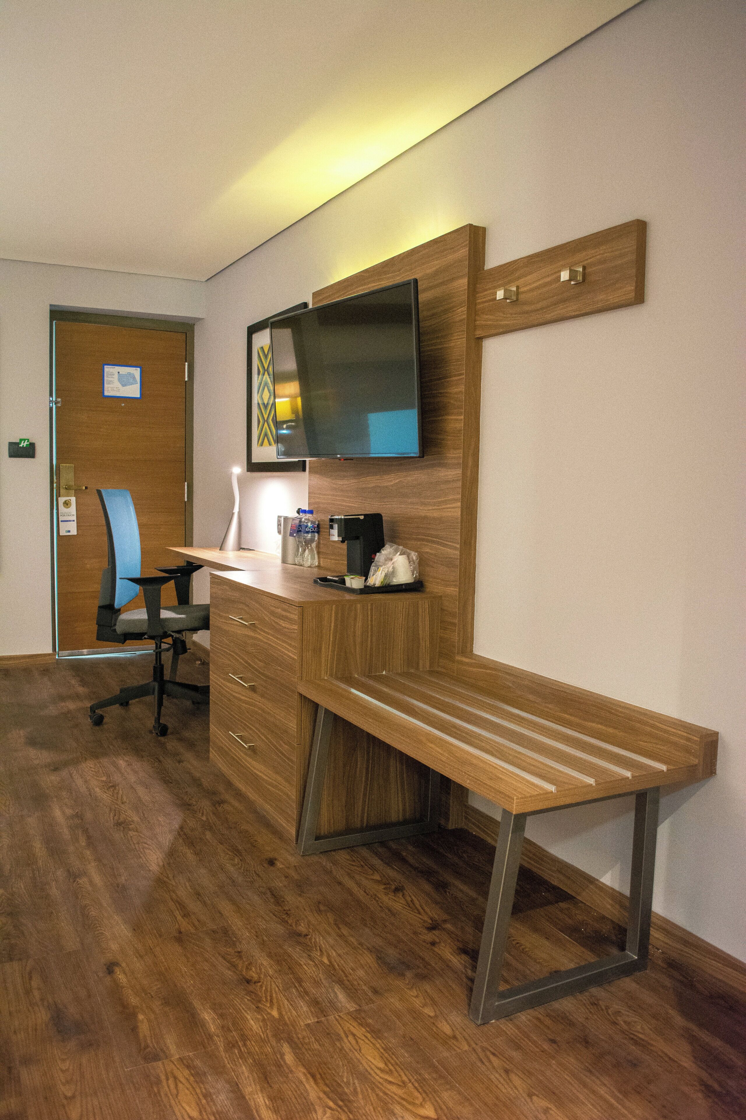 Quarto de hotel com uma escrivaninha de madeira, uma cadeira, uma televisão de tela plana fixada na parede e uma porta com uma placa.