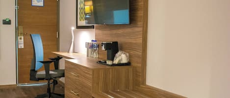 Quarto de hotel com uma escrivaninha de madeira, uma cadeira, uma televisão de tela plana fixada na parede e uma porta com uma placa.