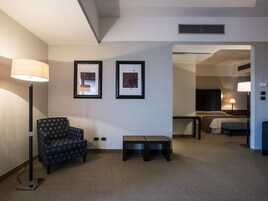 Quarto de hotel com uma cadeira, uma mesinha, duas fotografias emolduradas na parede, uma cama e um abajur.