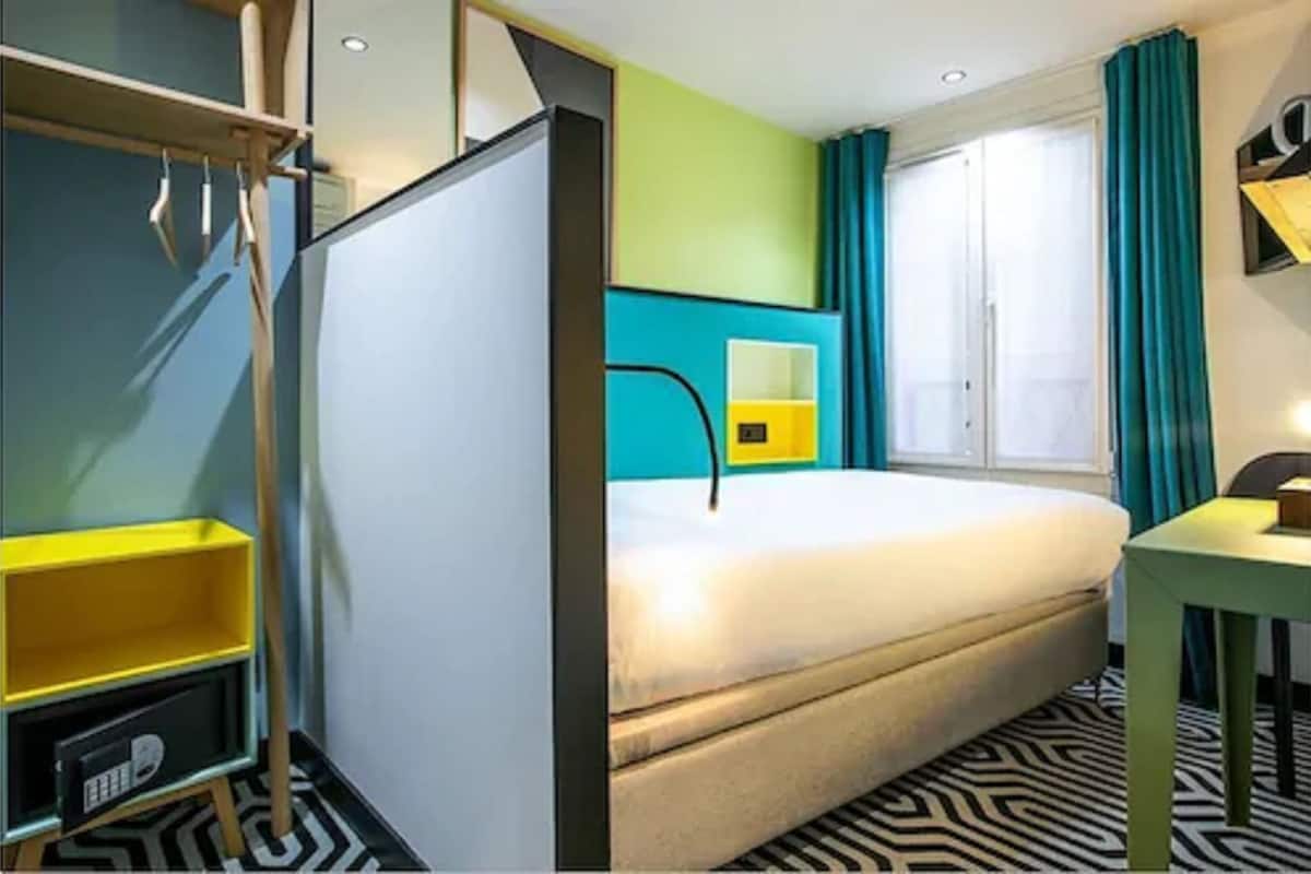 Quarto de hotel com cama, escrivaninha, cadeira, guarda-roupa e janela com cortinas.